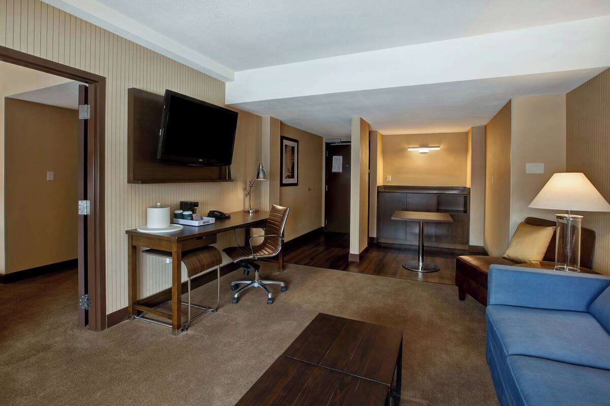 Quarto de hotel com escrivaninha, cadeira, televisão, sofá e uma pequena cozinha.
