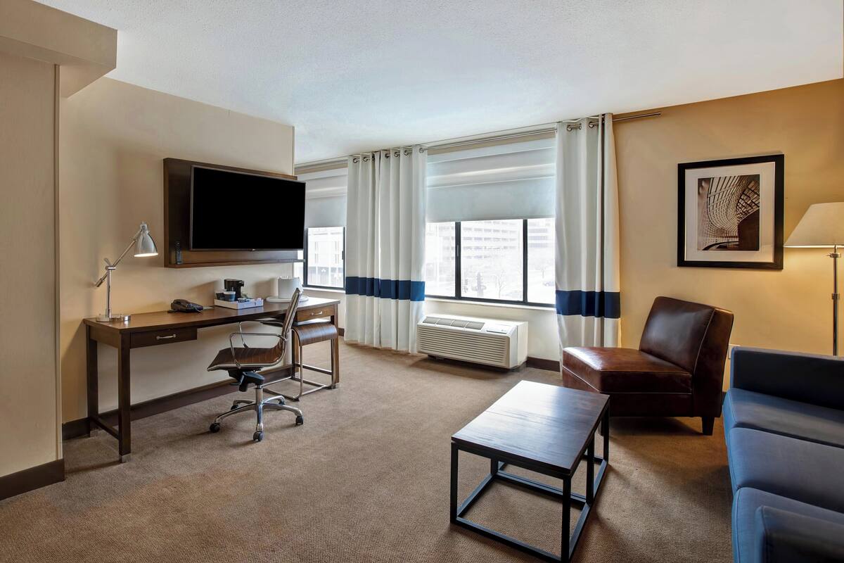 Quarto de hotel com escrivaninha, cadeira, televisão, sofá e um quadro na parede.