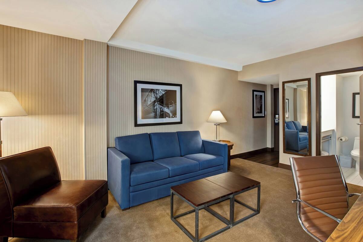 Quarto de hotel com sofá azul, poltrona de couro marrom, mesa de centro de madeira e quadro na parede.