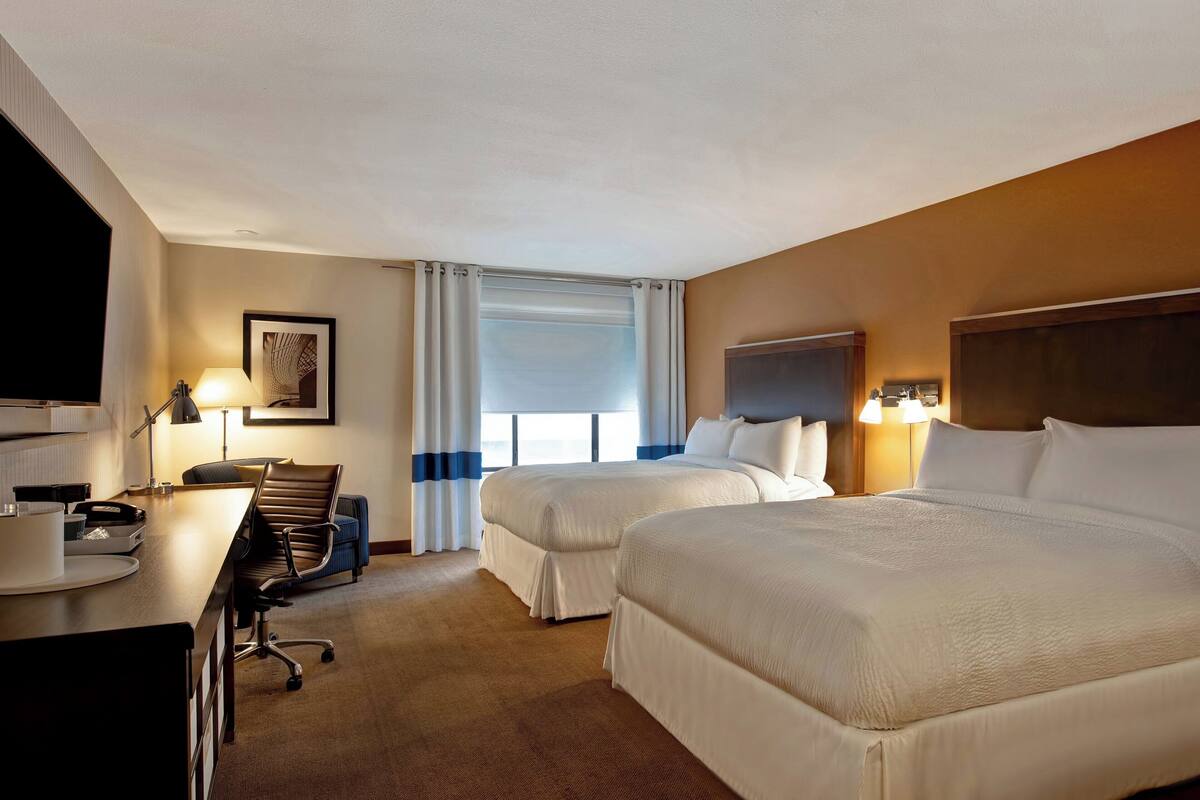 Quarto de hotel com duas camas, uma mesa, uma cadeira, uma televisão e uma janela com cortinas.