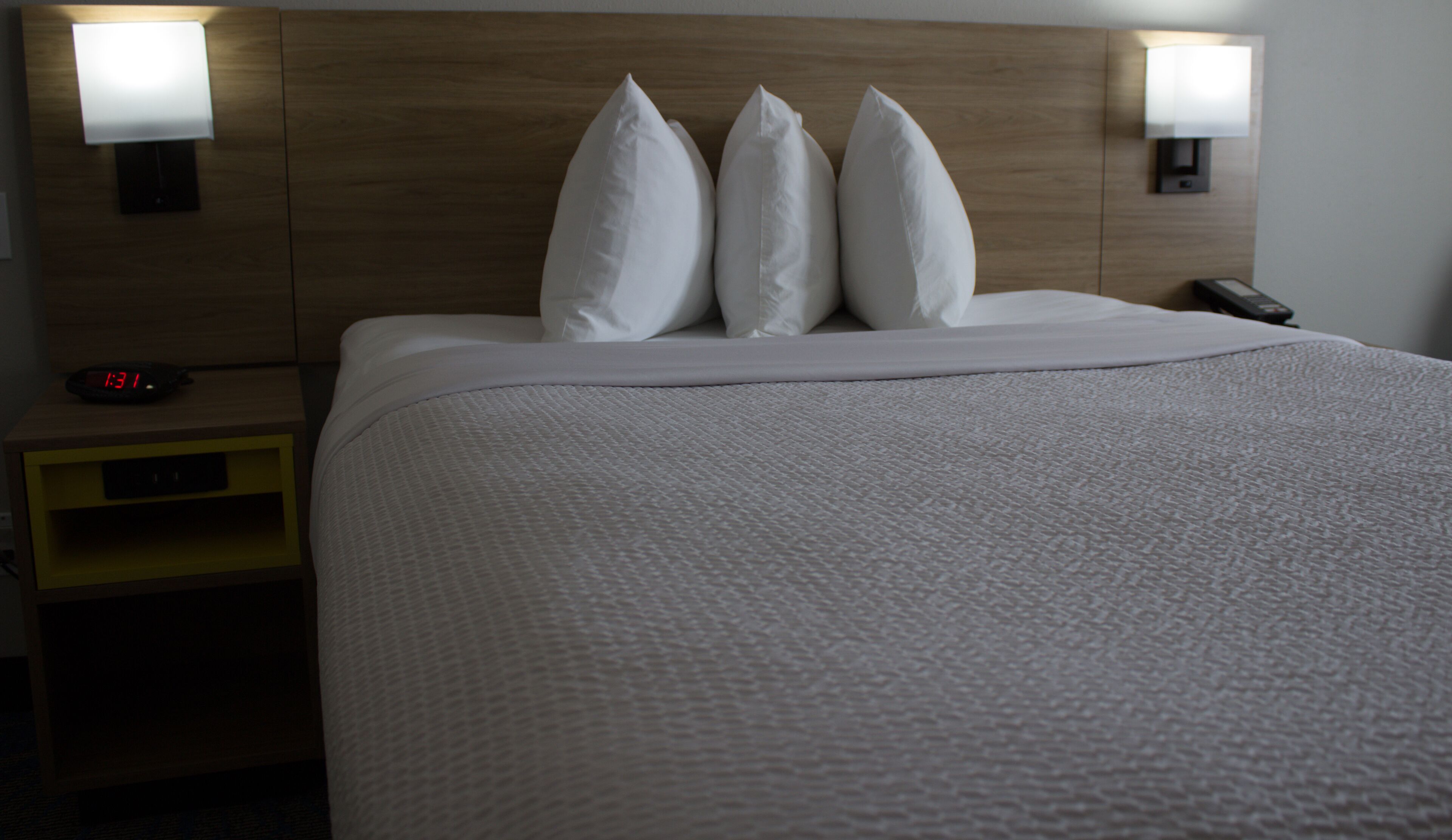 Quarto de hotel com cabeceira de madeira, cama com roupa de cama branca, duas luminárias de cabeceira, mesa de cabeceira com relógio digital e telefone.