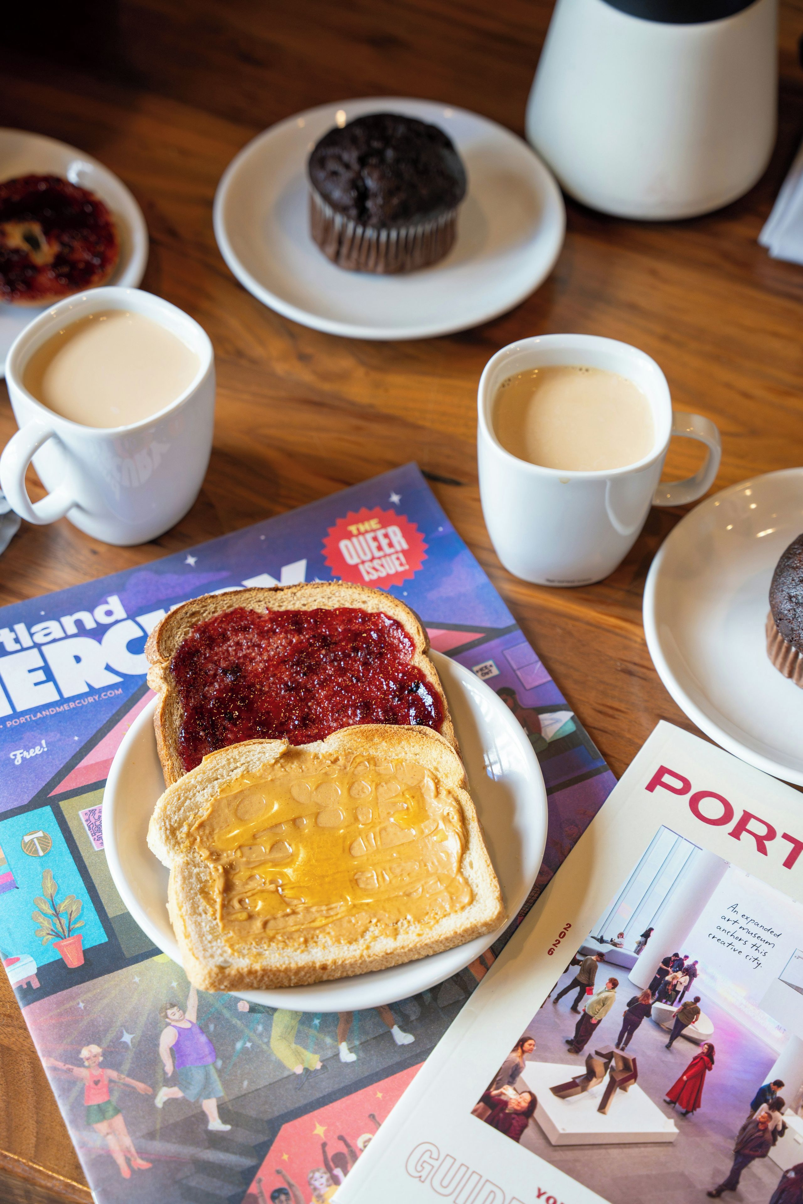 Uma mesa com um sanduíche, um muffin e uma xícara de café, junto com uma revista e um folheto.