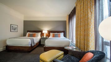 Quarto de hotel com duas camas, uma mesinha de cabeceira, uma luminária, uma cadeira e uma janela com cortinas.