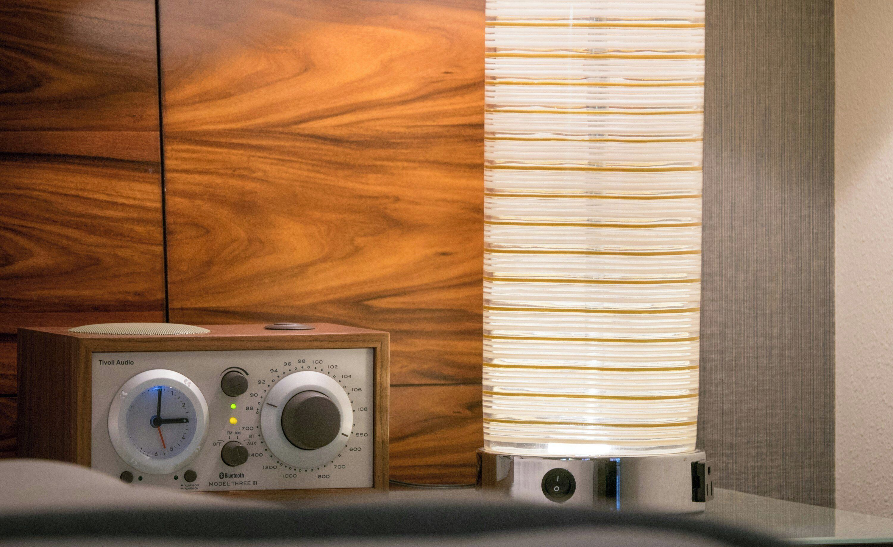 Um rádio com vários mostradores e um relógio, uma luminária com listras horizontais e uma parede de madeira.