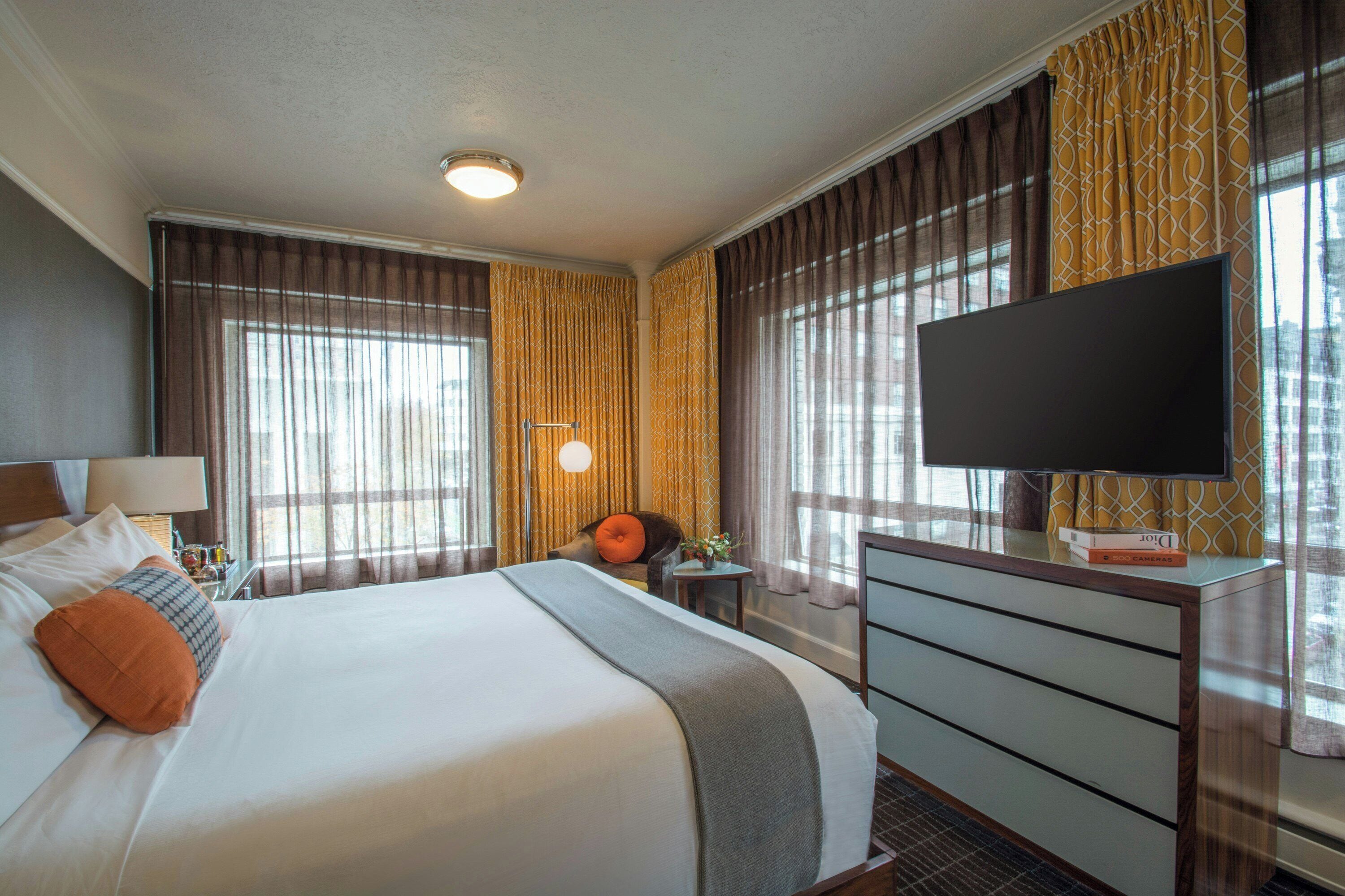 Quarto de hotel com cama, televisão, escrivaninha e janela com cortinas.