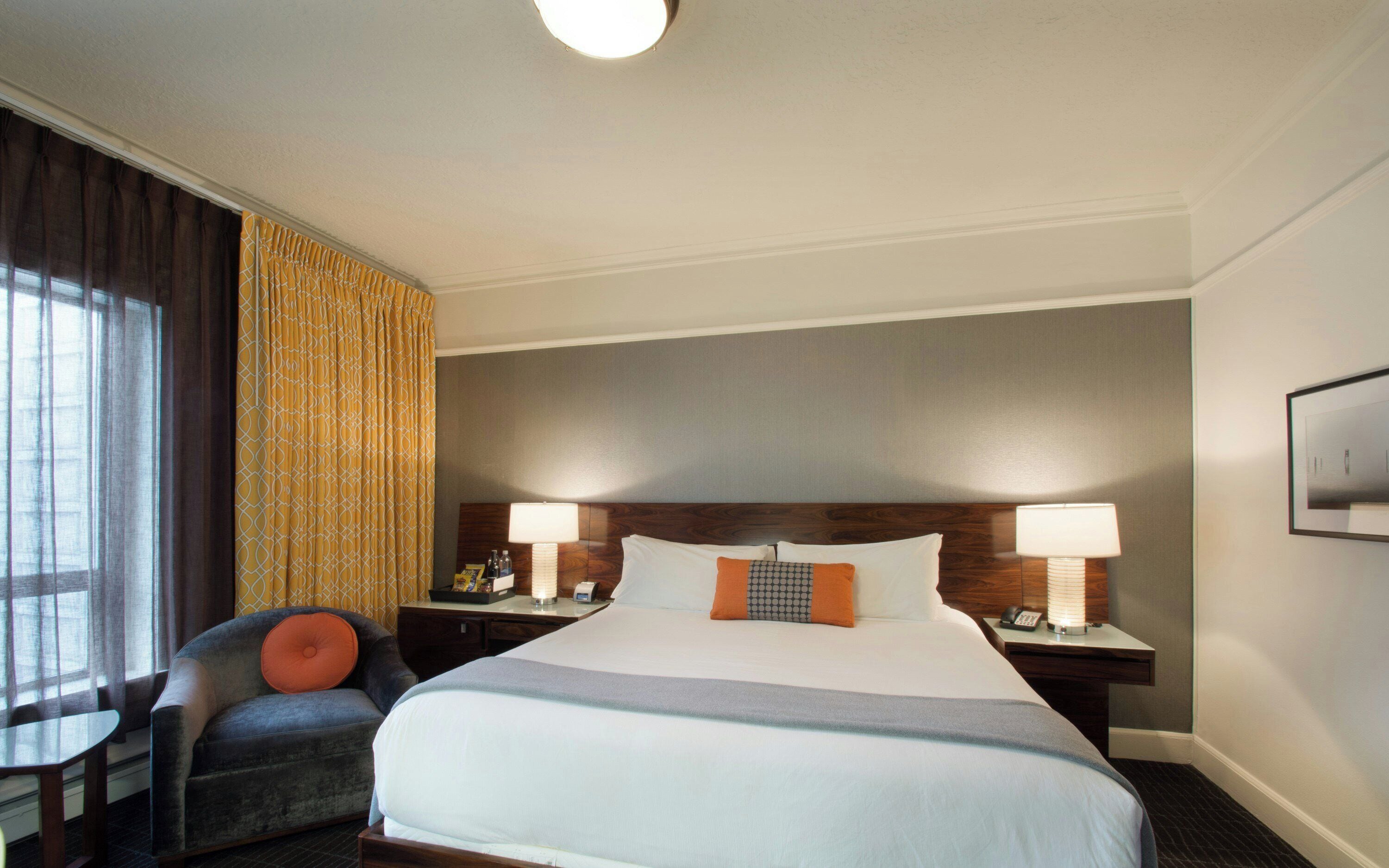 Quarto de hotel com uma cama grande, duas mesas de cabeceira com abajures, uma cadeira e uma janela com cortinas.