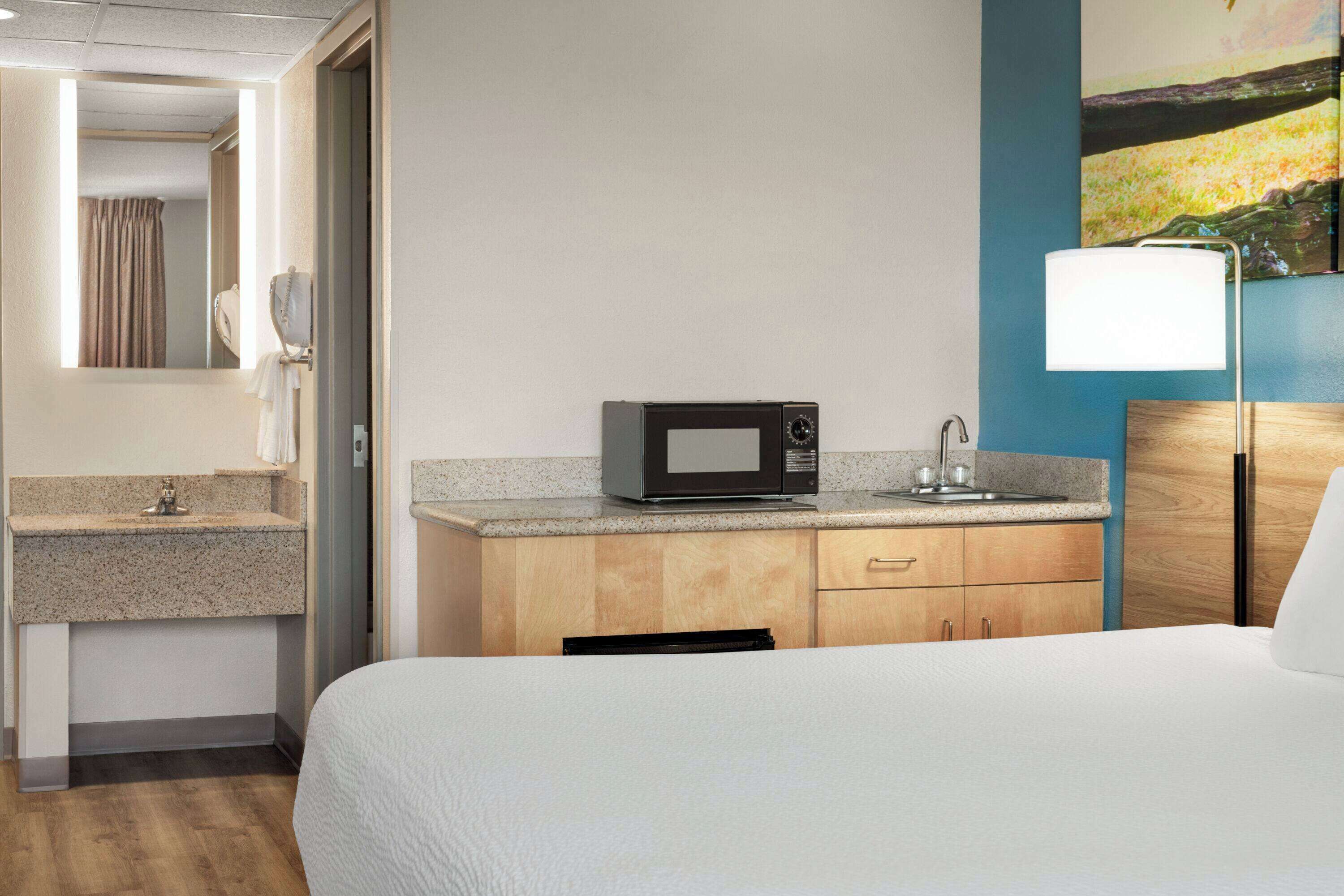Quarto de hotel com cama, micro-ondas na bancada, pia e espelho.