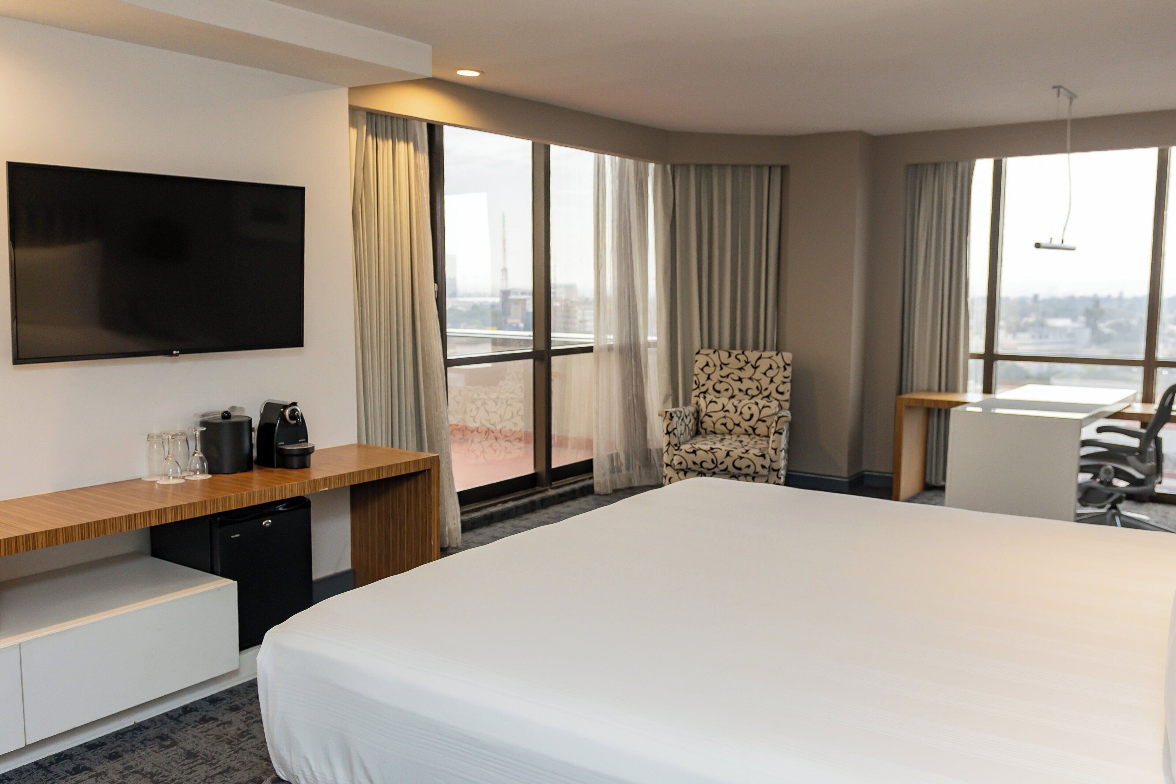 Quarto de hotel com uma cama grande, televisão de tela plana na parede, escrivaninha com cafeteira e uma cadeira com almofada estampada.