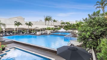Uma grande piscina ao ar livre com espreguiçadeiras e guarda-sóis, cercada por palmeiras e um edifício com telhado inclinado.