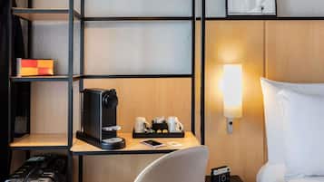 Quarto de hotel com cama, escrivaninha com cafeteira, cadeira e prateleira com uma caixa e uma caneca.