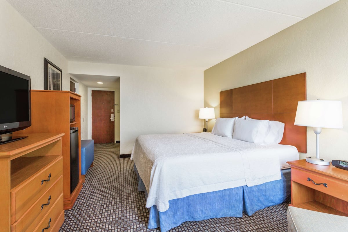 Quarto de hotel com cama, luminárias de cabeceira, televisão, cómoda de madeira e banheiro com chuveiro e vaso sanitário.