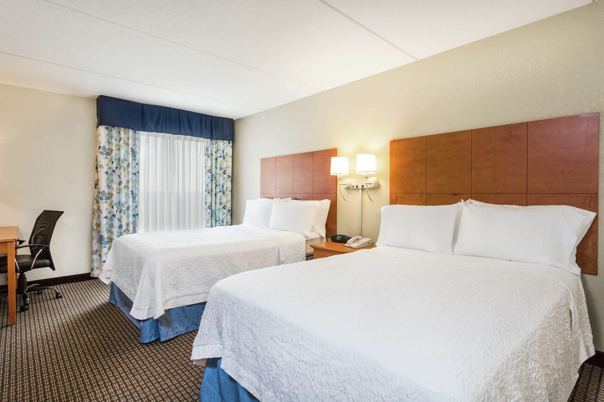 Quarto de hotel com duas camas, uma escrivaninha, uma cadeira e uma mesa de cabeceira com abajur.