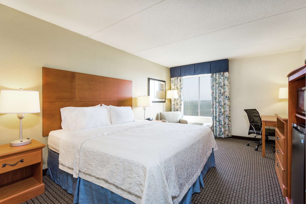 Quarto de hotel com uma cama grande, uma escrivaninha, uma cadeira, uma televisão e uma janela com cortinas.