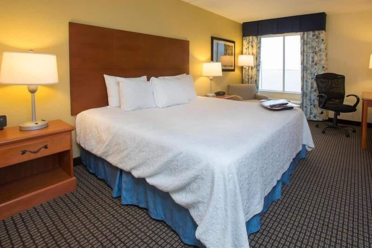 Quarto de hotel com cama, abajures, escrivaninha, cadeira e janela com cortinas.
