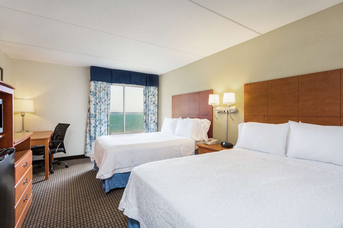 Quarto de hotel com duas camas, uma escrivaninha, uma cadeira, um armário e uma janela com cortinas.