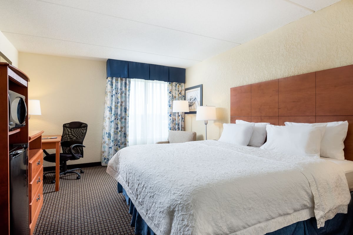 Quarto de hotel com uma cama grande, uma escrivaninha com cadeira, cômoda e janela com cortinas.