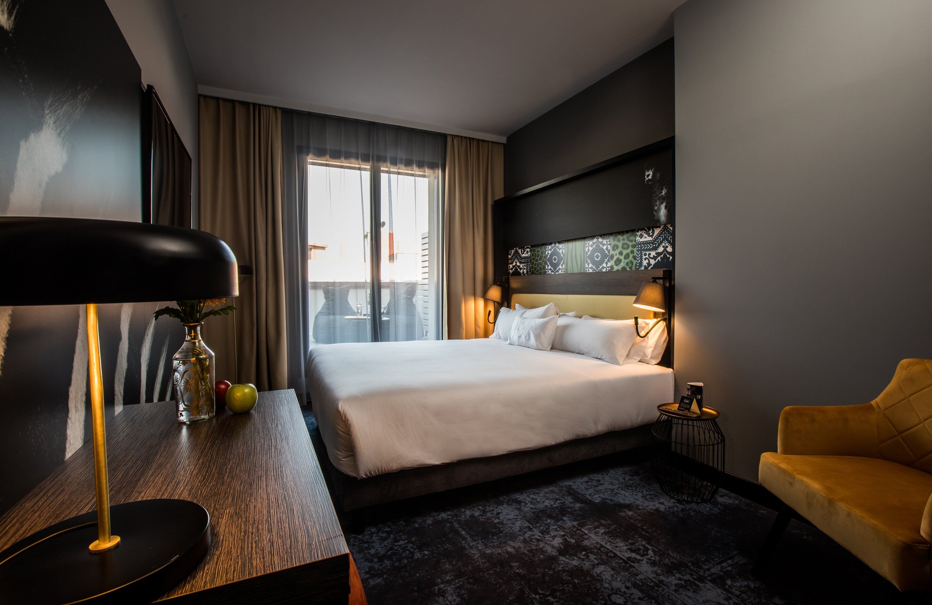 Quarto de hotel com cama, mesa de cabeceira, abajur e uma cadeira.