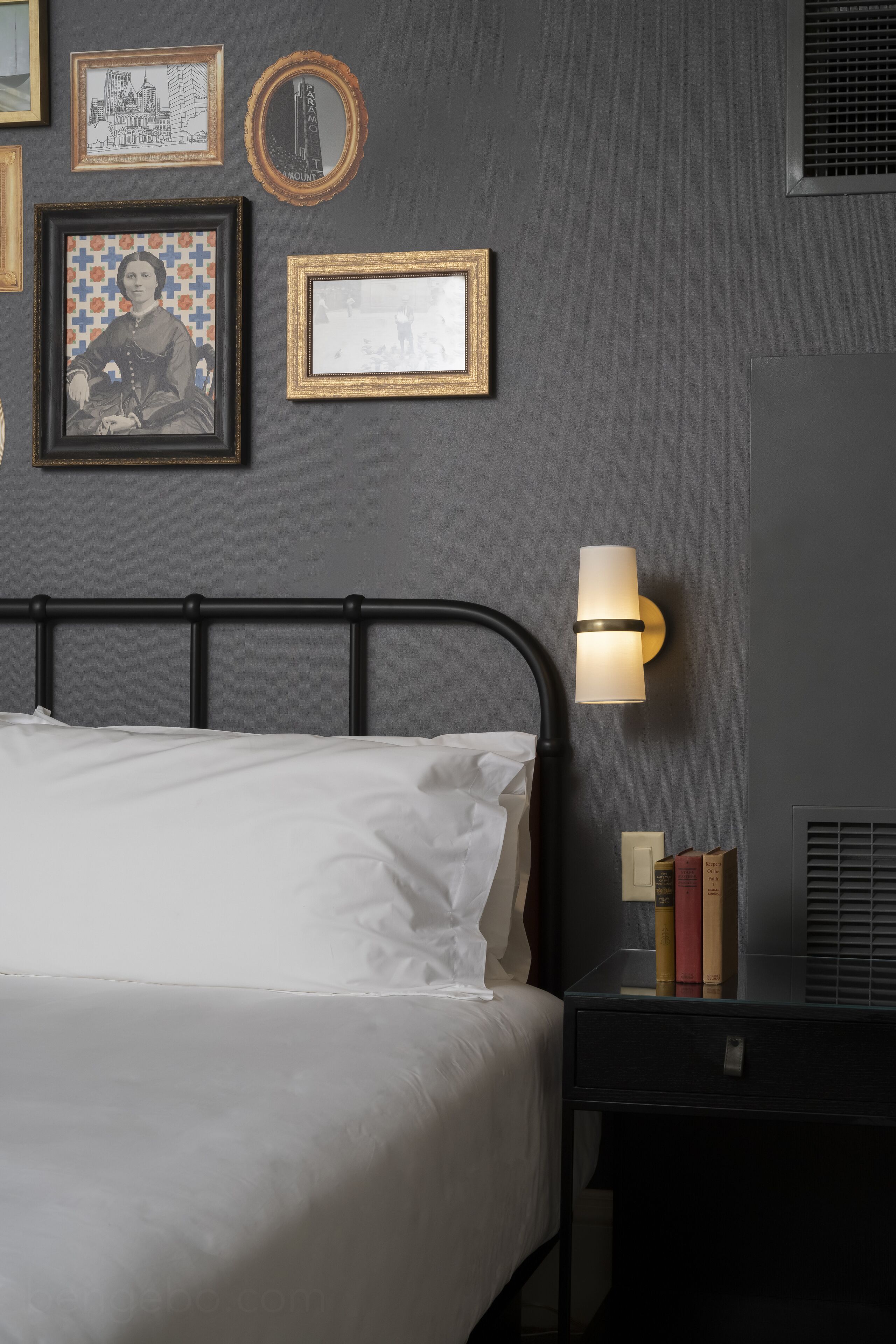Uma cama bem arrumada com lençóis brancos, cabeceira de metal preto e mesa de cabeceira com abajur e livros.