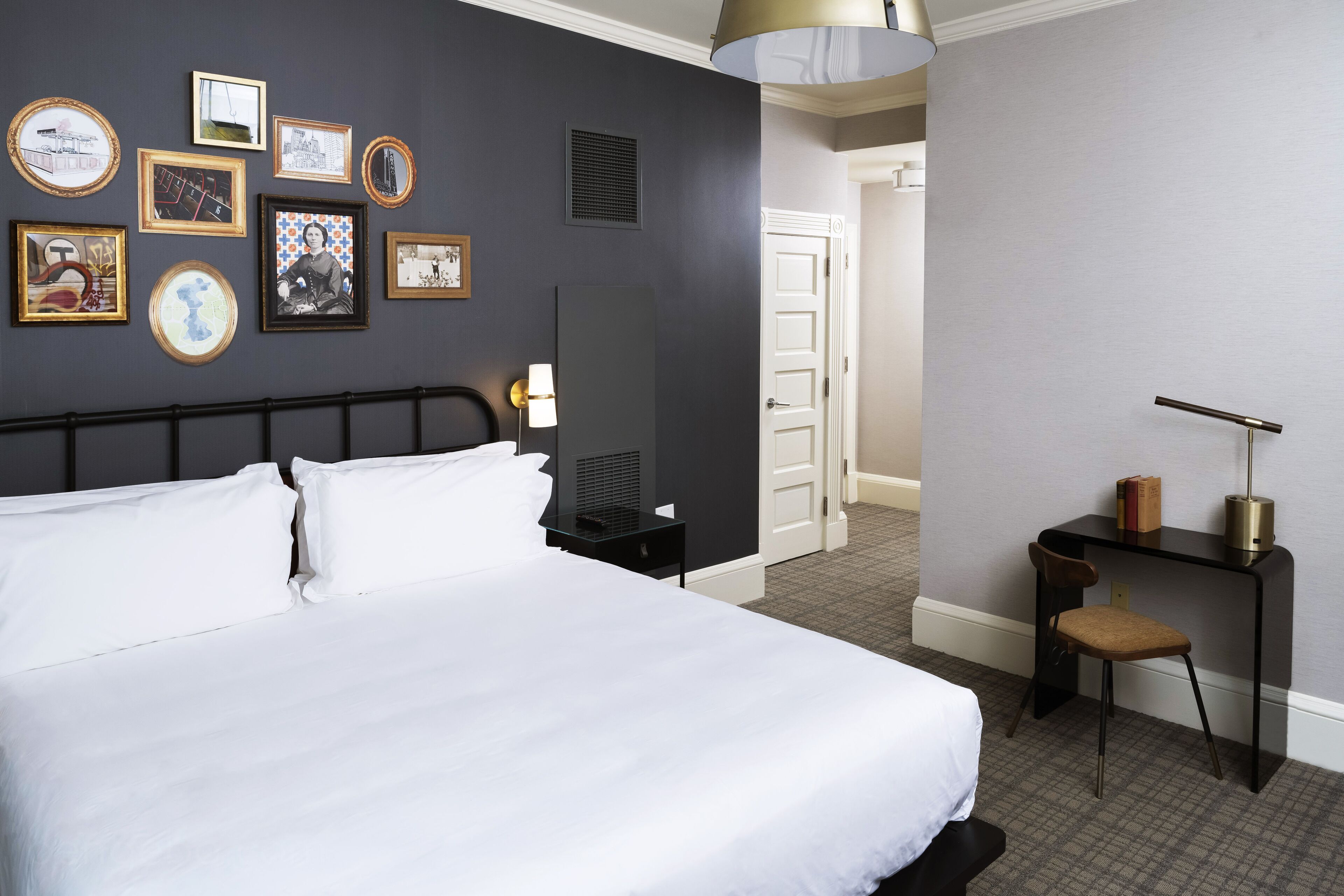 Quarto de hotel com uma cama grande, uma mesa de trabalho com cadeira e parede decorada com quadros.