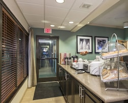 Corredor de hotel com sinalização de saída, área de kitchenette com máquinas de café e buffet com pães e doces.
