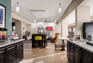 Um saguão de hotel moderno com balcão de recepção, caixa eletrônico, área de refeições com cadeiras e cozinha com geladeira.