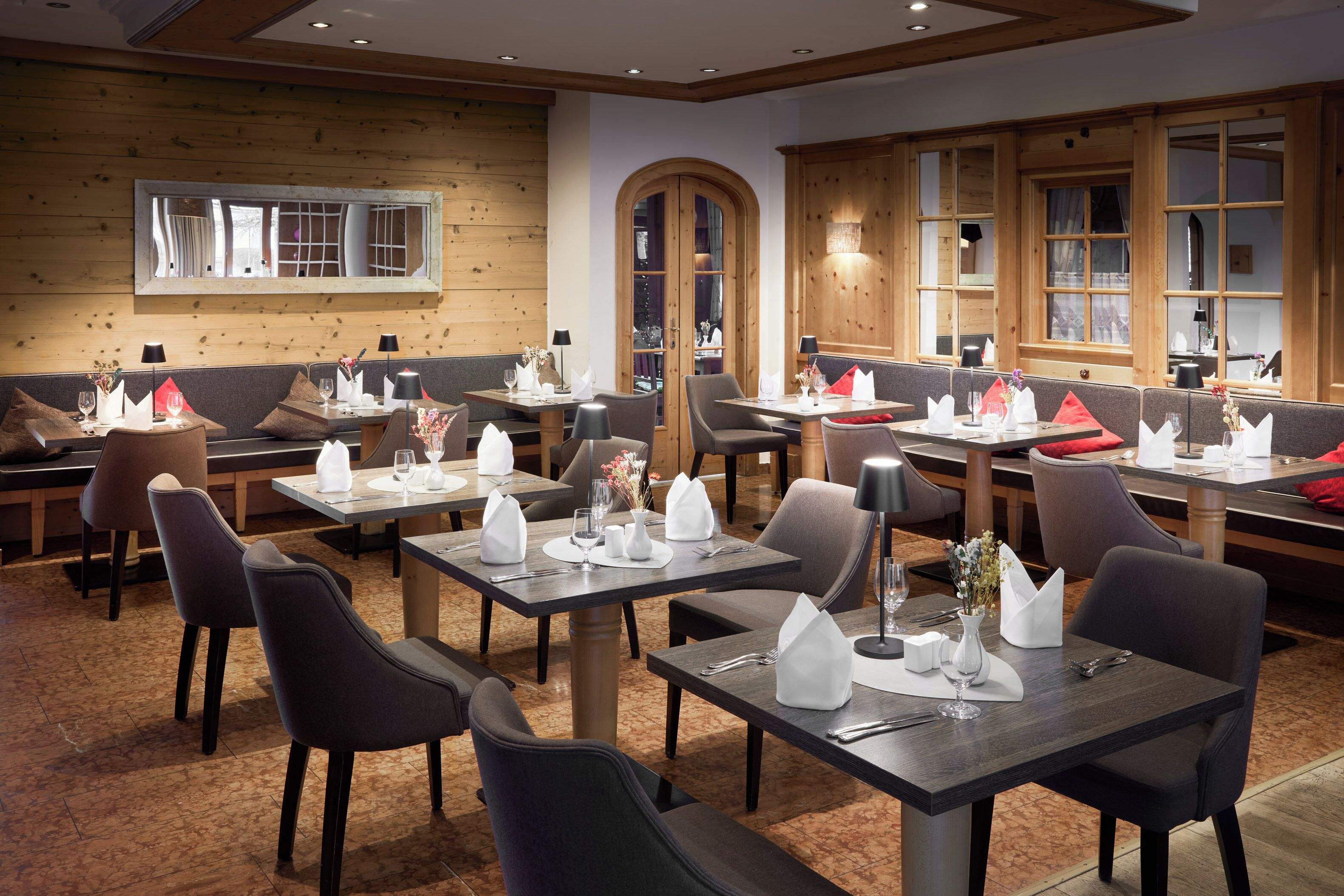 Restaurante com paredes de madeira e grandes janelas, mesas com guardanapos brancos e talheres, e uma combinação de bancos e cadeiras.