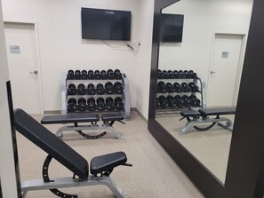 Academia com equipamentos de musculação, um banco e um espelho refletindo os aparelhos.