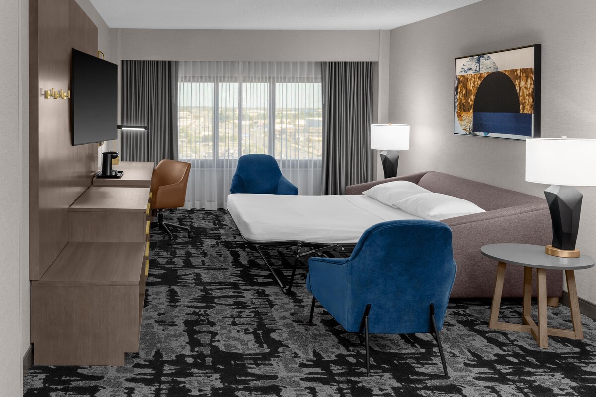 Quarto de hotel com cama, duas cadeiras, escrivaninha com televisão, abajur e um quadro na parede.