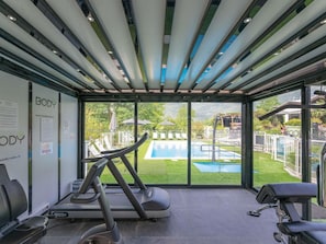 Uma academia com equipamentos de ginástica, piscina e vista para uma área ajardinada.