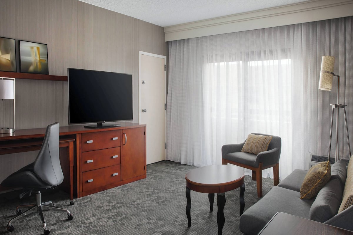 Quarto de hotel com televisão de tela plana, uma escrivaninha com gavetas, cadeira de escritório cinza, sofá cinza, mesa redonda pequena e abajur.