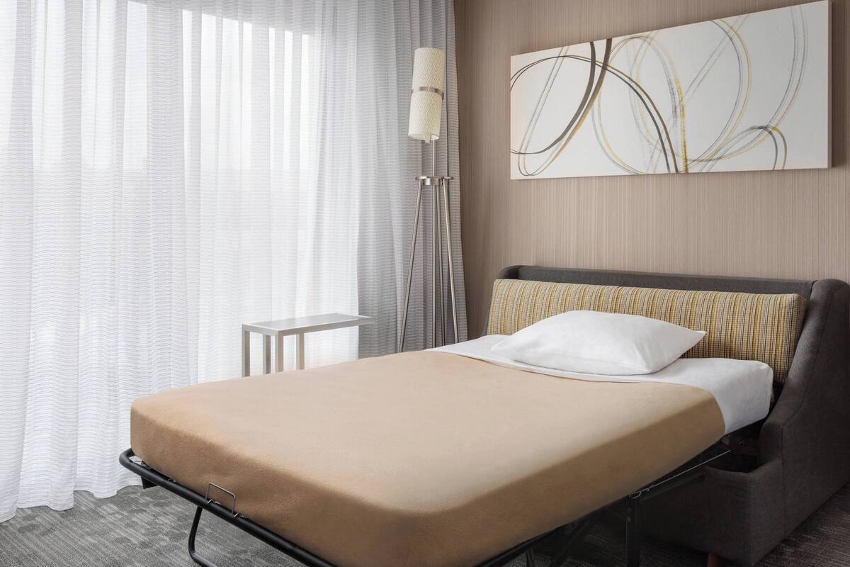Quarto de hotel moderno com uma cama, um abajur, uma mesinha e um quadro abstrato grande na parede.