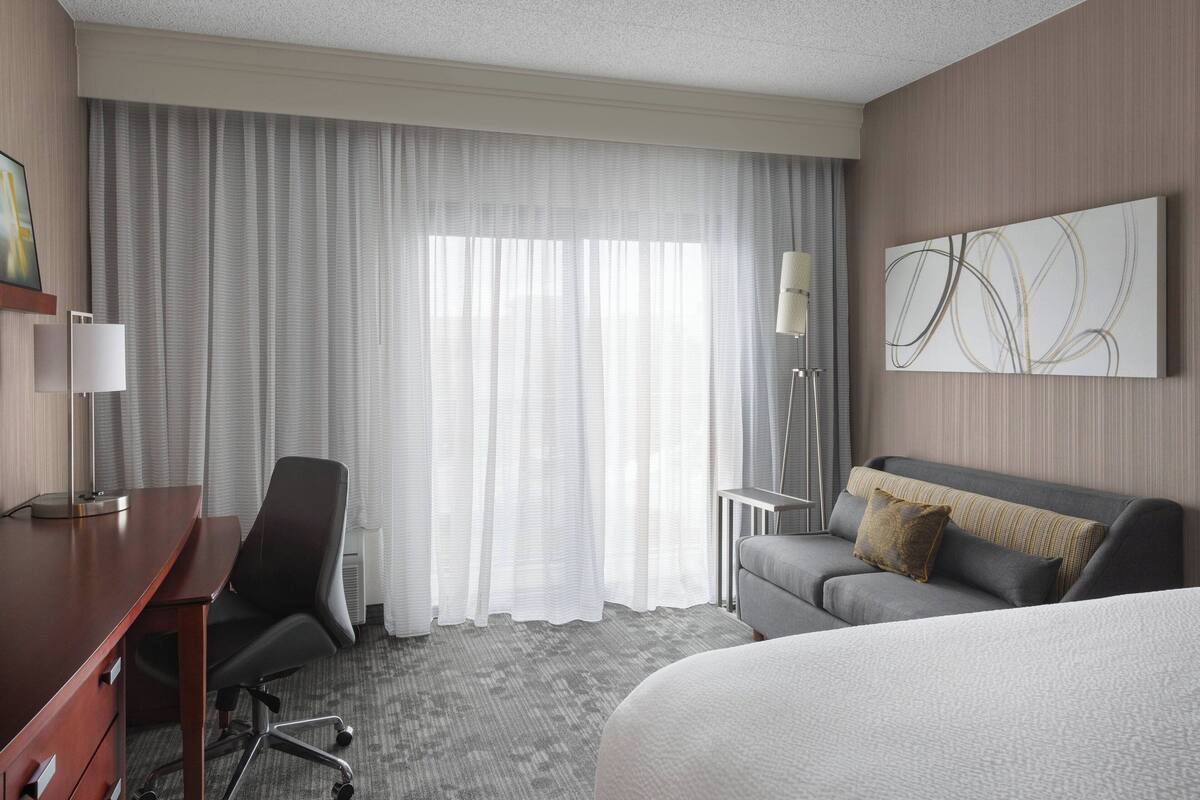 Quarto de hotel com uma cama, uma mesa, uma cadeira, um sofá e uma janela grande com cortinas.