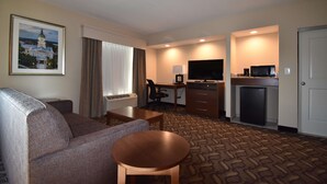 Quarto de hotel com sofá, mesa redonda pequena, escrivaninha com computador e televisão, mini-geladeira e uma porta.