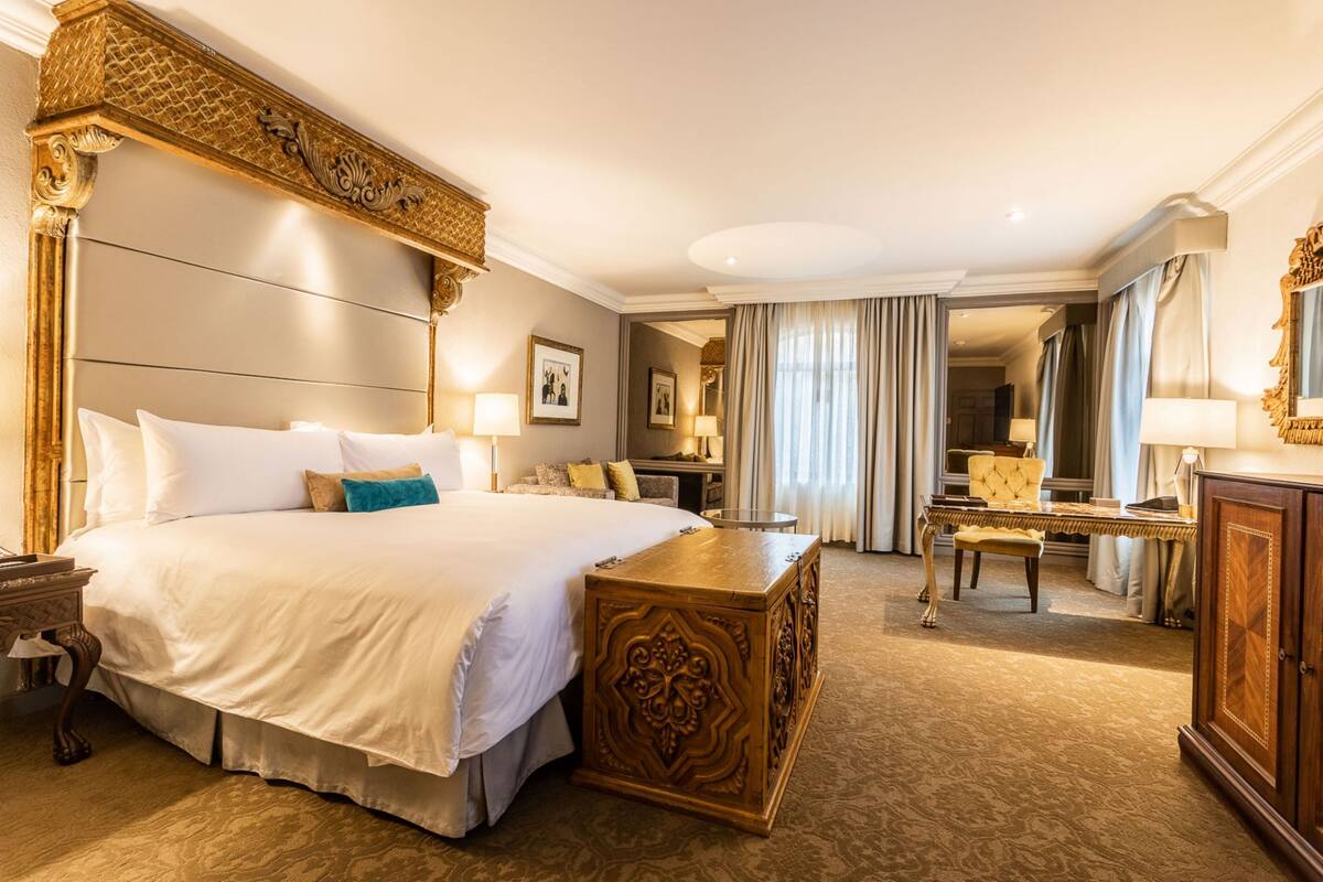 Quarto de hotel espaçoso com uma cama grande, uma mesa de cabeceira de madeira, uma escrivaninha e um guarda-roupa.
