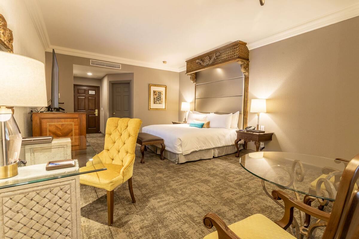 Quarto de hotel com cama, mesa de vidro, cadeira, escrivaninha e espelho.