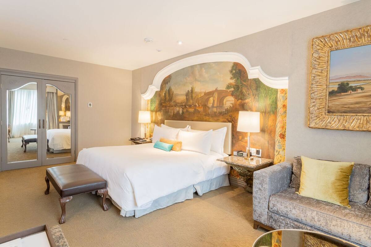 Quarto de hotel com uma cama grande, um espaço de estar com sofá, uma mesinha e um espelho.