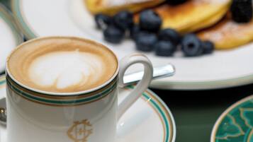 Uma chávena de café com arte de latte num pires, um prato com panquecas, mirtilos e amoras, e uma pequena chávena de chá.
