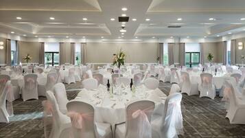 Um amplo salão de banquetes com mesas redondas preparadas para refeições, cadeiras brancas com fitas cor-de-rosa e grandes janelas com cortinas.