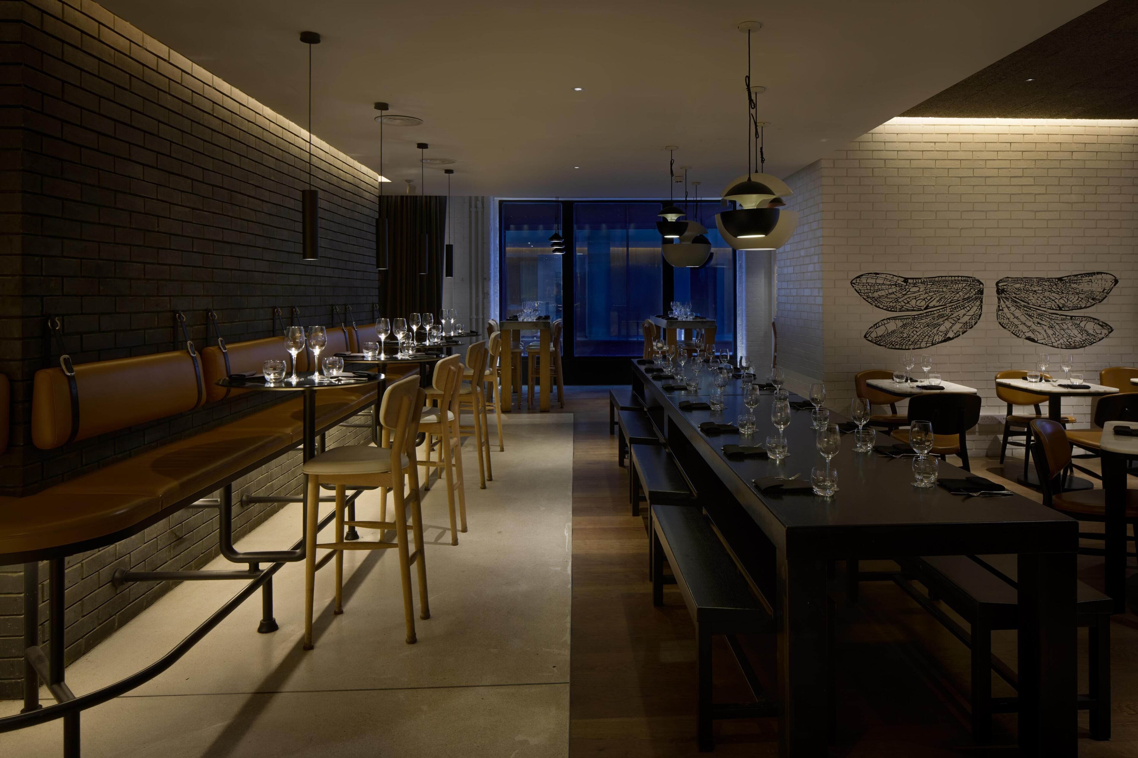 Um restaurante com mesas grandes e compartilhadas, cadeiras de madeira e luminárias pendentes.