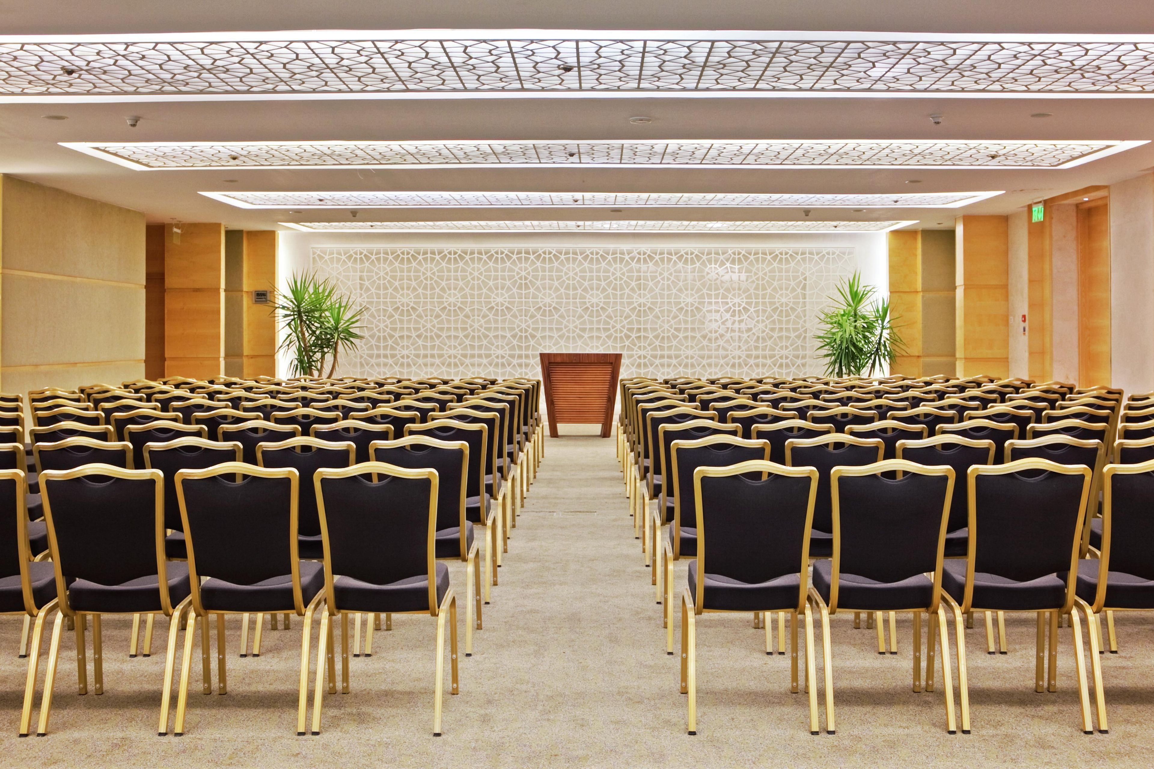 Sala de conferências com fileiras de cadeiras, um púlpito e painéis decorativos na parede.