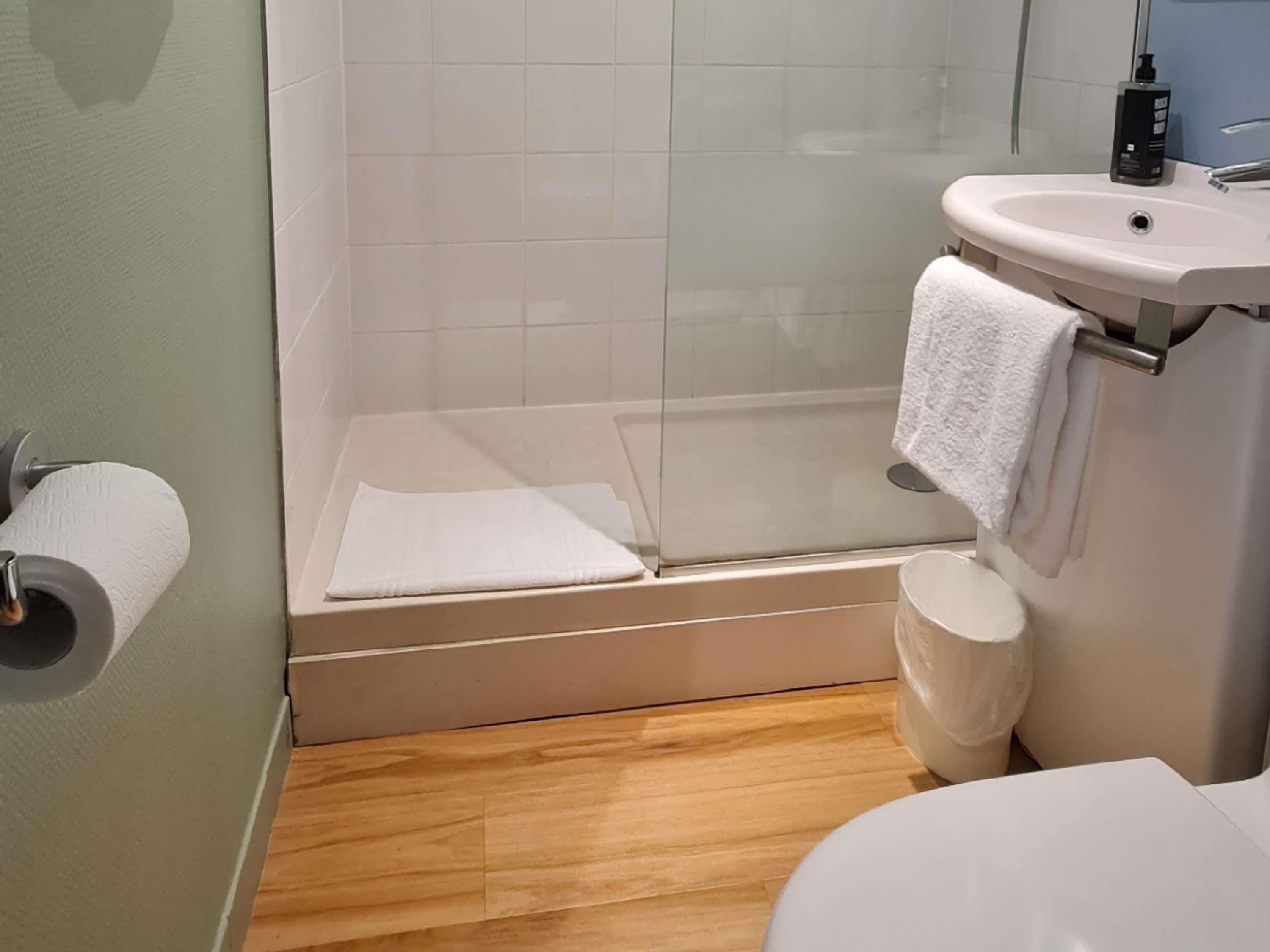 Casa de banho com sanita, lavatório, cabine de duche em vidro e rolo de papel higiénico.