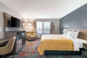 Quarto de hotel com cama, escrivaninha, cadeira, televisão e janela com cortinas.