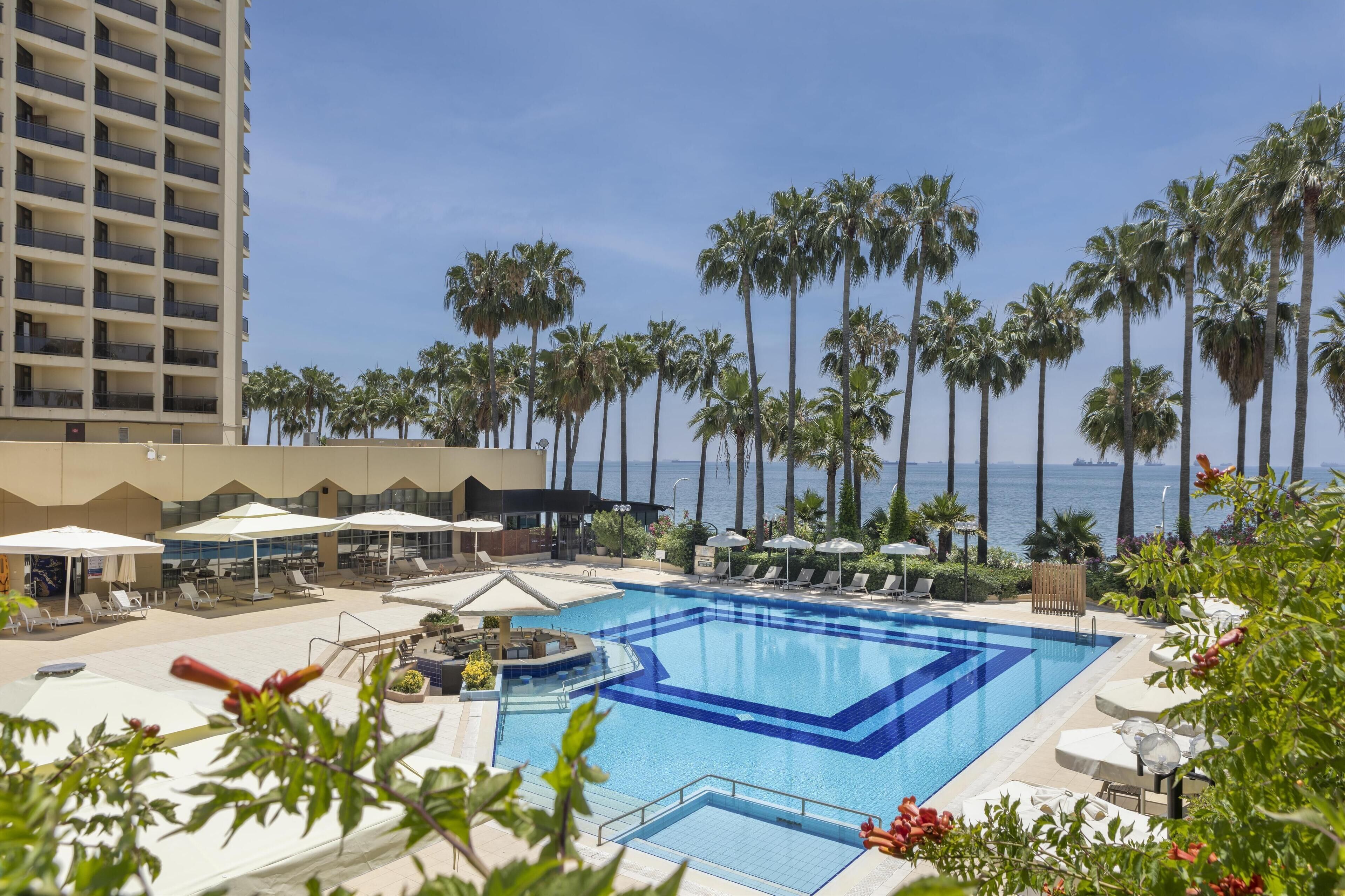 Área de piscina de hotel com vista para o oceano, cercada por palmeiras e espreguiçadeiras.