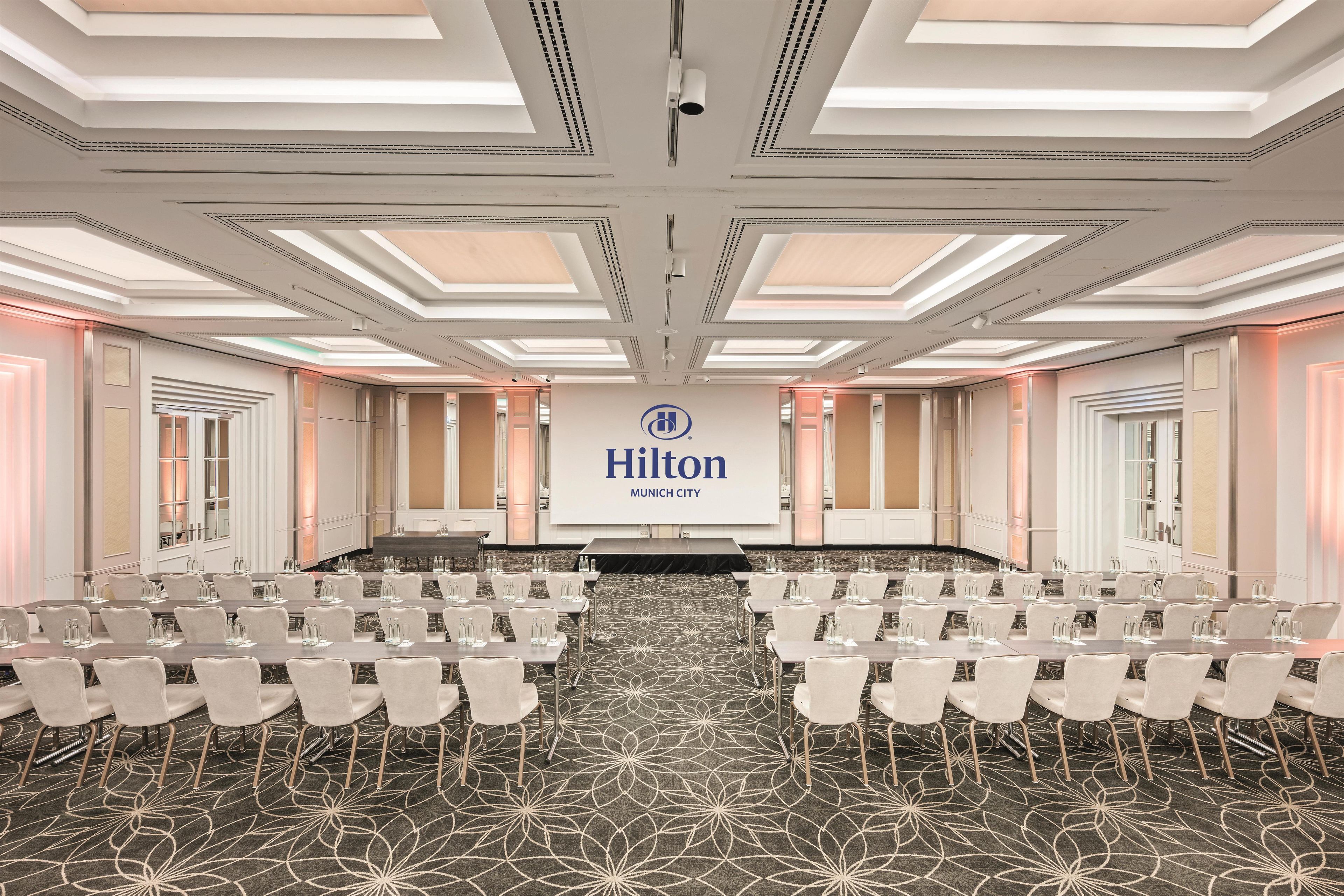 Uma sala de conferências com fileiras de cadeiras, um palco e uma tela grande exibindo 'Hilton Munich City'.