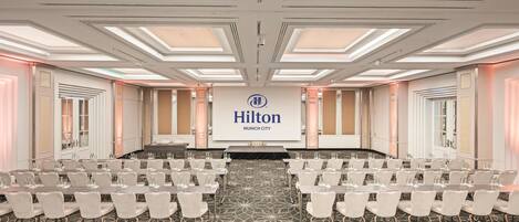 Uma sala de conferências com fileiras de cadeiras, um palco e uma tela grande exibindo 'Hilton Munich City'.