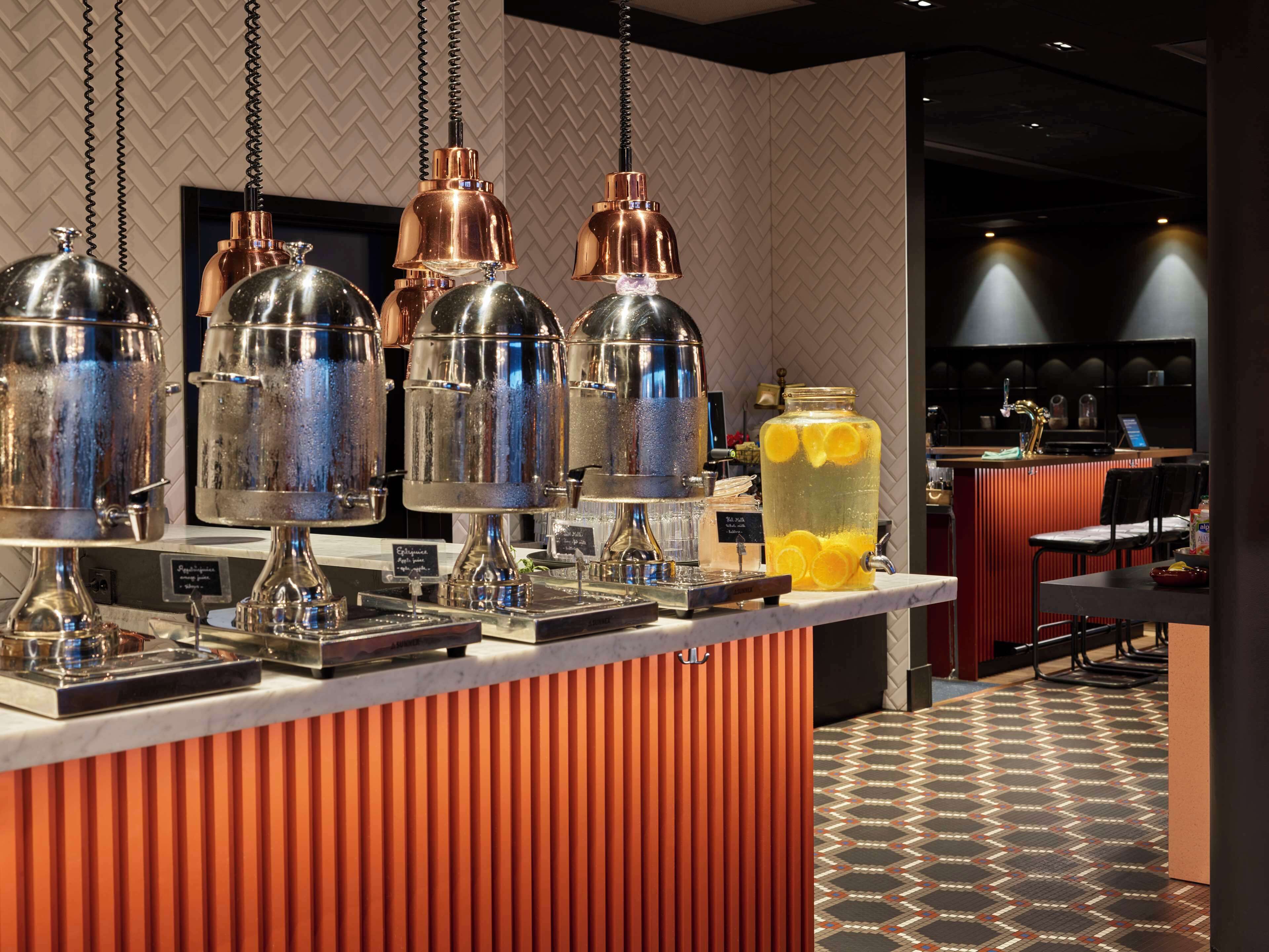 Uma área de bar com dosadores de bebidas em cobre e vidro, um grande pote de vidro com fatias de cítricos e um chão estampado.