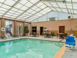 Uma piscina interior com espreguiçadeiras e uma mesa, rodeada por paredes de tijolo e um teto de vidro.