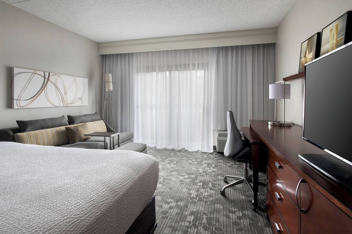 Quarto de hotel com uma cama grande, uma escrivaninha, uma cadeira, uma TV e uma janela com cortinas.