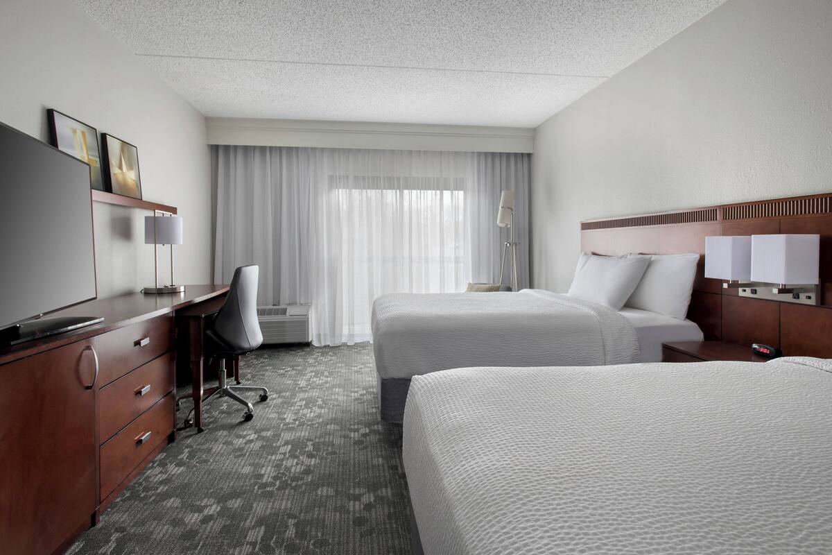 Quarto de hotel com duas camas, uma escrivaninha, uma cadeira e uma janela grande com cortinas.