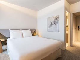 Quarto de hotel com uma cama grande, cabeceira, mesa de cabeceira, obra de arte na parede e uma porta que dá para outro cômodo.